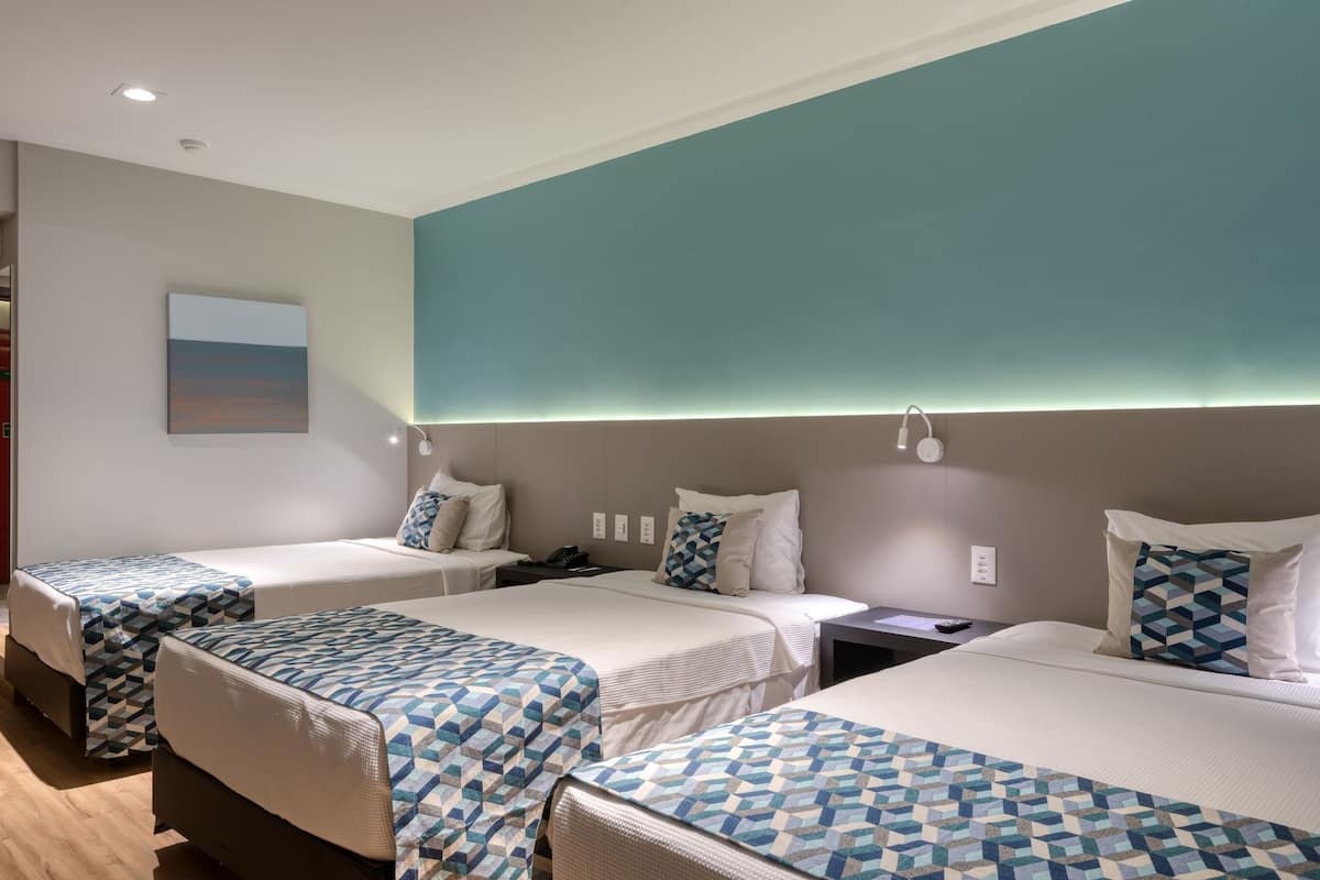Quarto de hotel com três camas, cada uma com colcha estampada em padrão geométrico. Há um quadro na parede, uma mesa de cabeceira com telefone e luminária fixa.