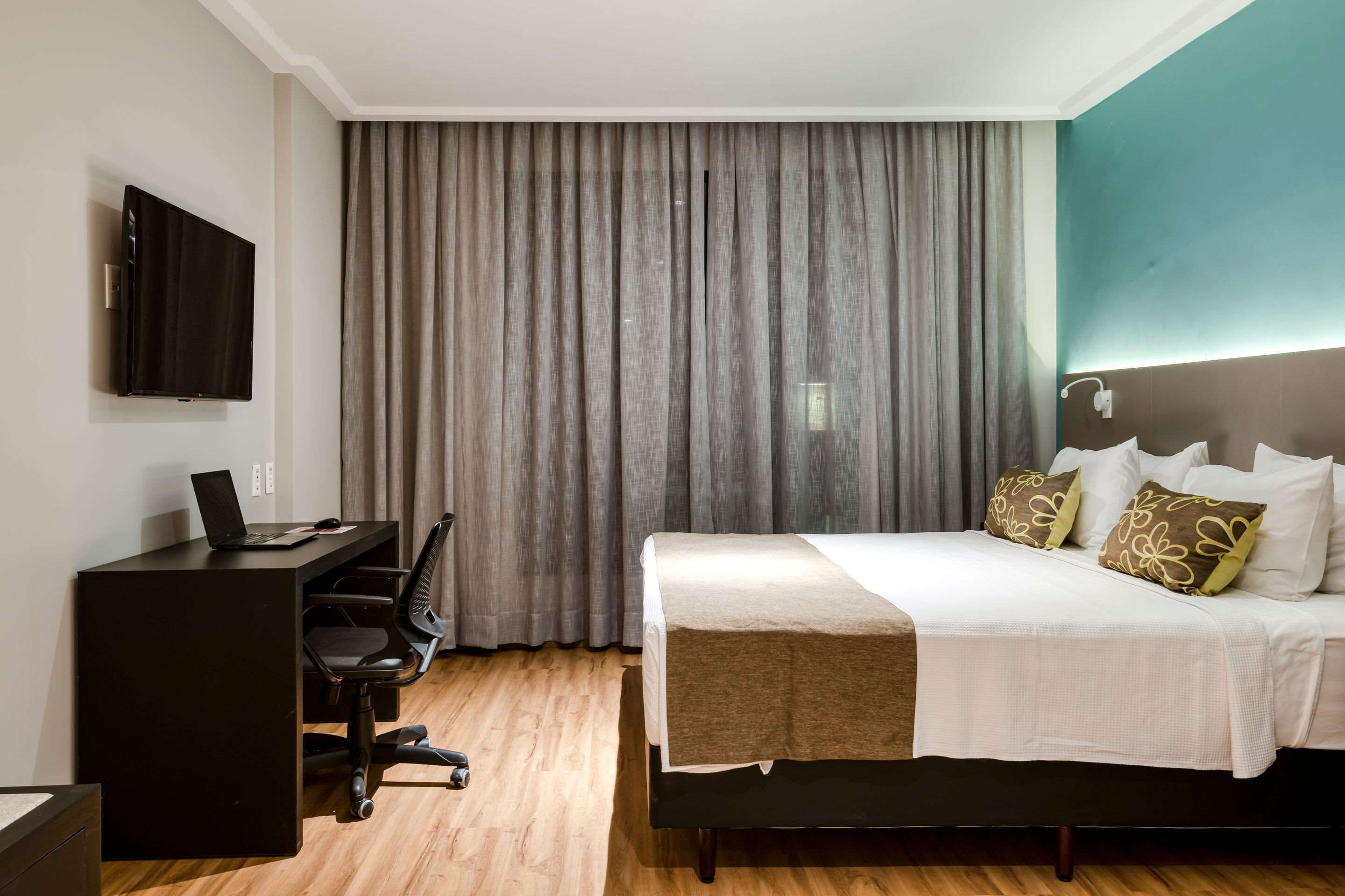 Quarto de hotel com cama, escrivaninha com laptop, televisão e cortina.