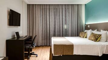 Quarto de hotel com cama, escrivaninha com laptop, televisão e cortina.