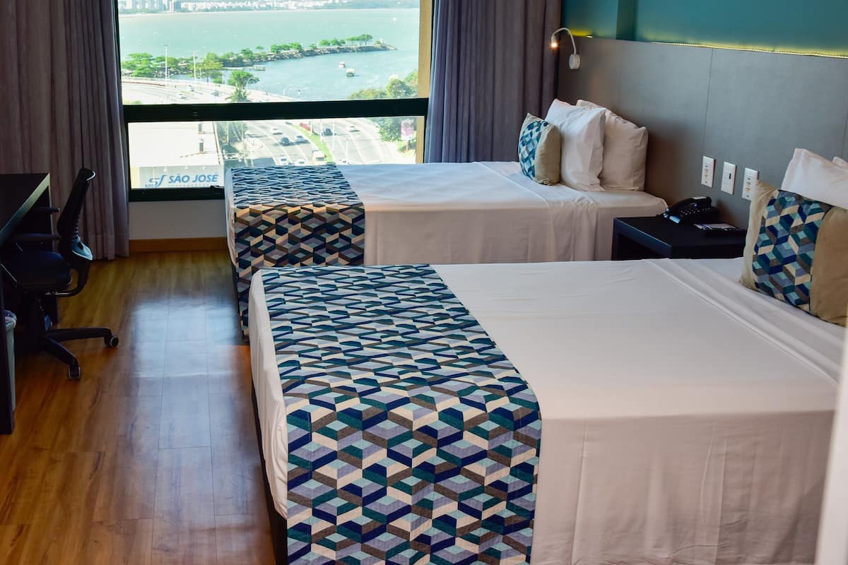 Quarto de hotel com duas camas, uma escrivaninha e vista para um corpo de água.