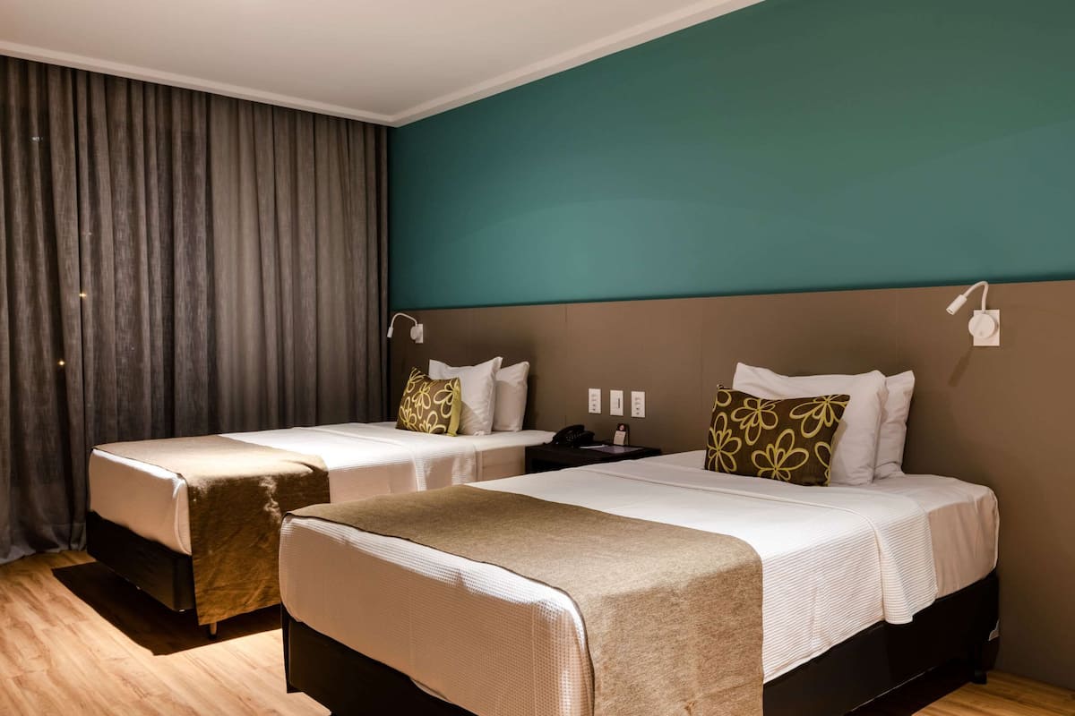 Quarto de hotel com duas camas, uma janela grande com cortinas, uma parede de destaque azul-turquesa e piso de madeira.