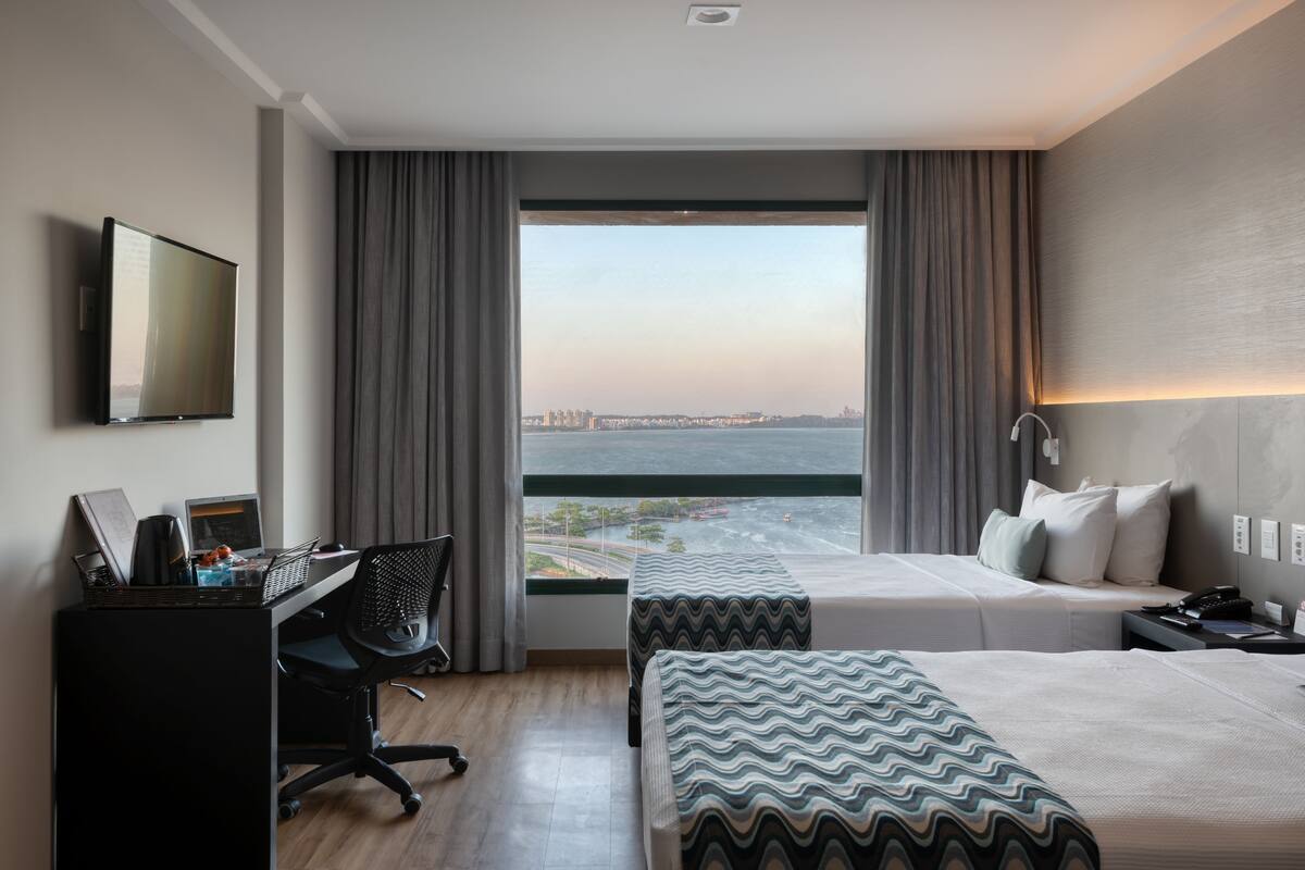 Quarto de hotel com escrivaninha, duas camas, televisão e vista para a cidade e para a água.
