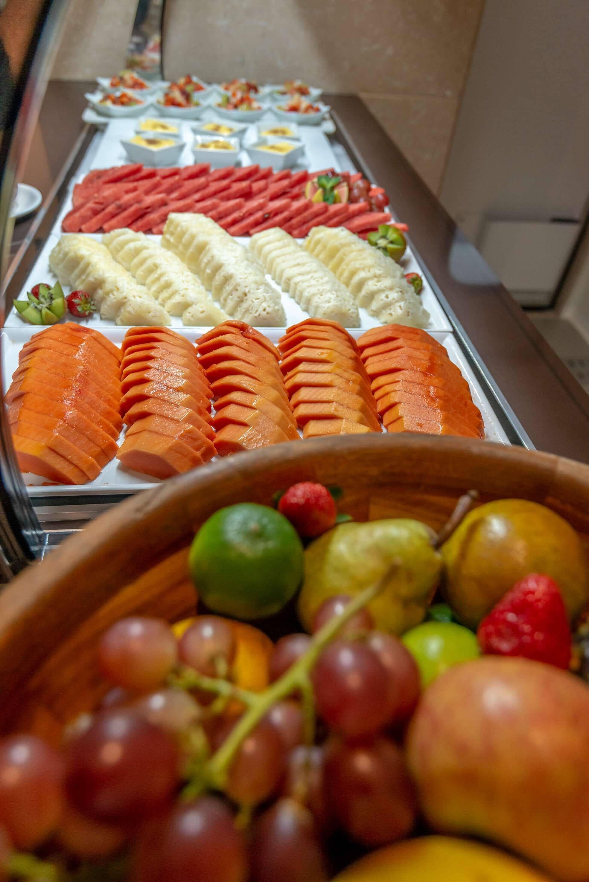 Vitrine com frutas e legumes fatiados, incluindo melancia, bananas e uvas.
