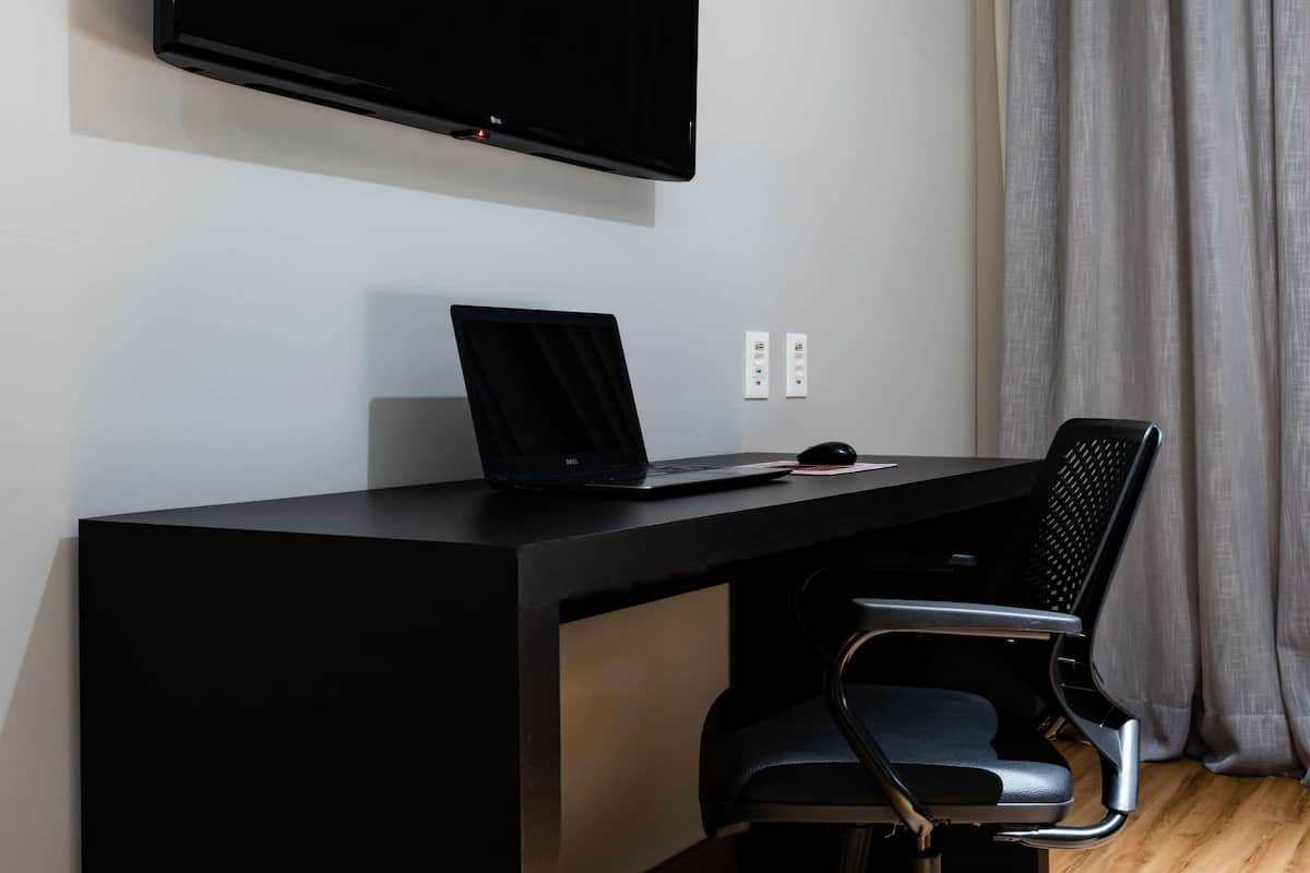 Um espaço de escritório moderno com uma mesa preta, um laptop, uma cadeira, uma televisão presa à parede e uma janela com cortinas.