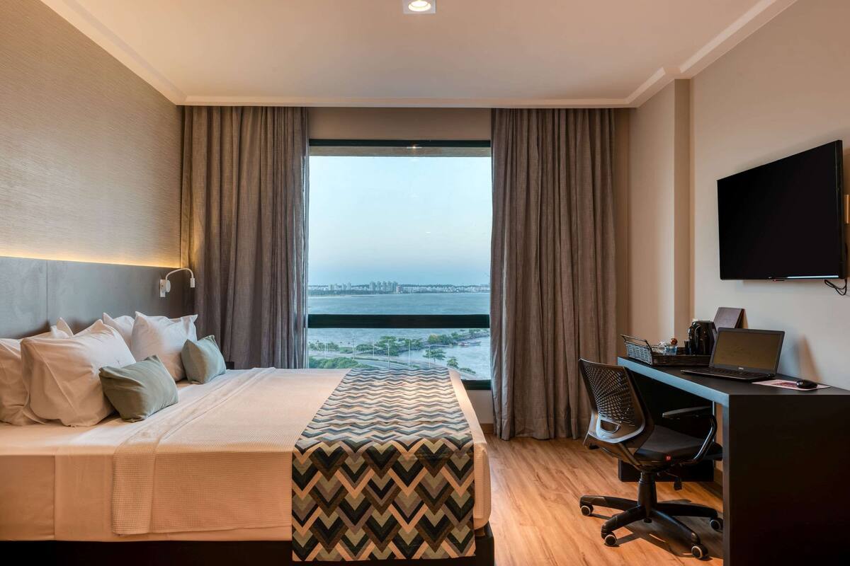 Quarto de hotel com cama, escrivaninha, laptop, cadeira e televisão.
