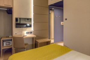Quarto de hotel com cama, escrivaninha, cadeira, televisão e guarda-roupa.