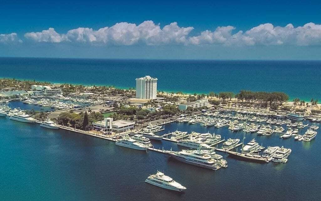Vista aérea de uma marina com vários barcos atracados, um hotel imponente e uma vista desobstruída do oceano.
