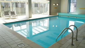 Piscina interna com paredes de vidro transparente, chão revestido com azulejos e algumas cadeiras de descanso.