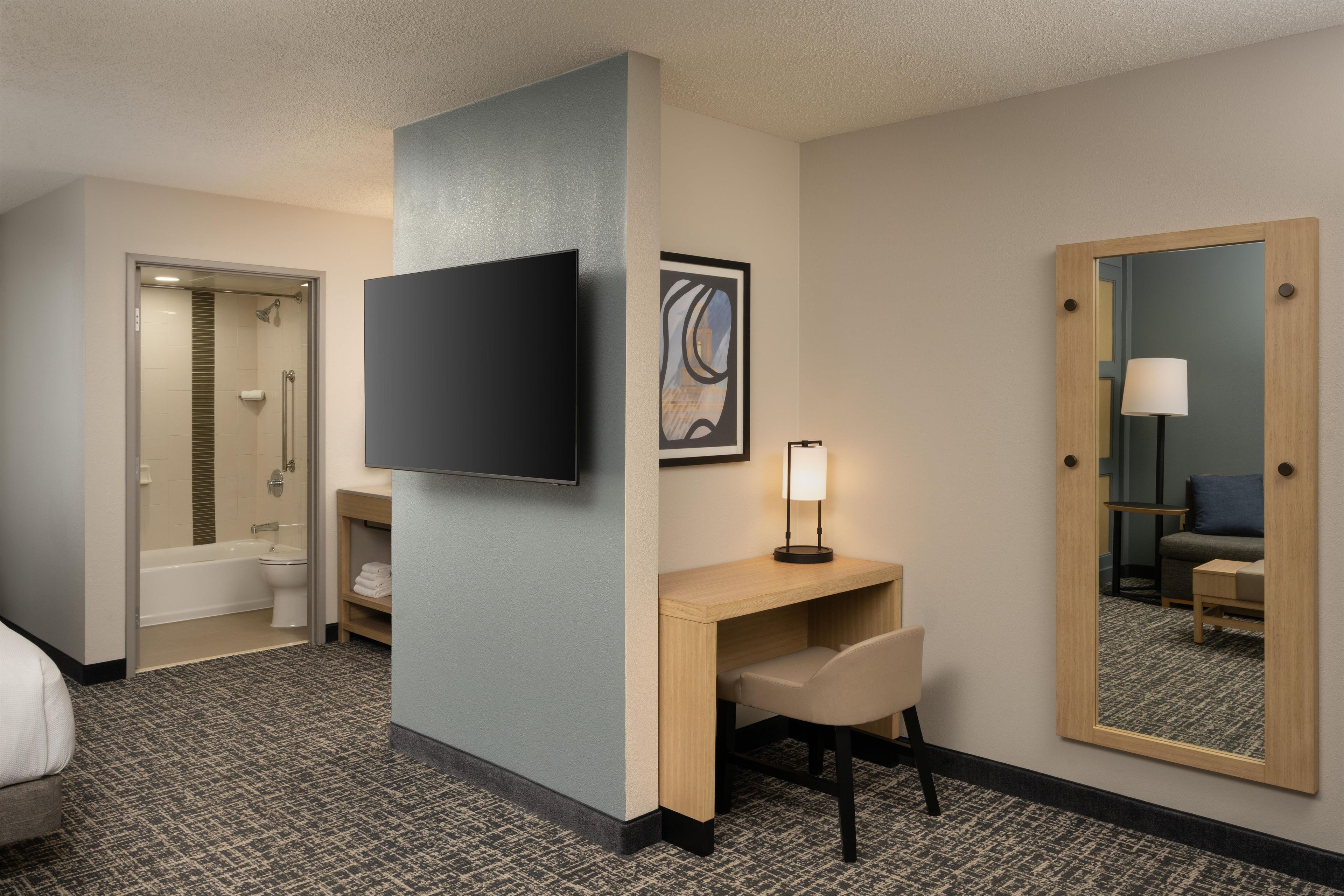 Quarto de hotel com televisão de tela plana na parede, escrivaninha com cadeira, espelho grande, área de chuveiro e uma cama.