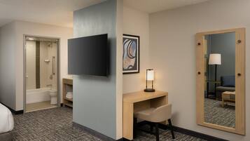 Quarto de hotel com televisão de tela plana na parede, escrivaninha com cadeira, espelho grande, área de chuveiro e uma cama.