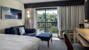 Quarto de hotel com cama, sofá, escrivaninha e vista para árvores pela janela.