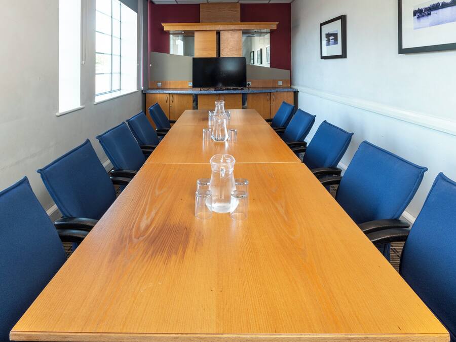 Uma sala de conferências com uma longa mesa de madeira, cadeiras azuis, uma TV sobre um suporte de madeira e quadros nas paredes.