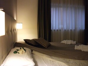 Quarto de hotel com uma cama, mesa de cabeceira, abajur e janela com cortinas.