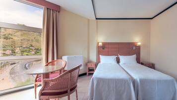 Quarto de hotel com cama, mesas de cabeceira, uma cadeira, uma mesinha e vista para a praia e casas pela janela.