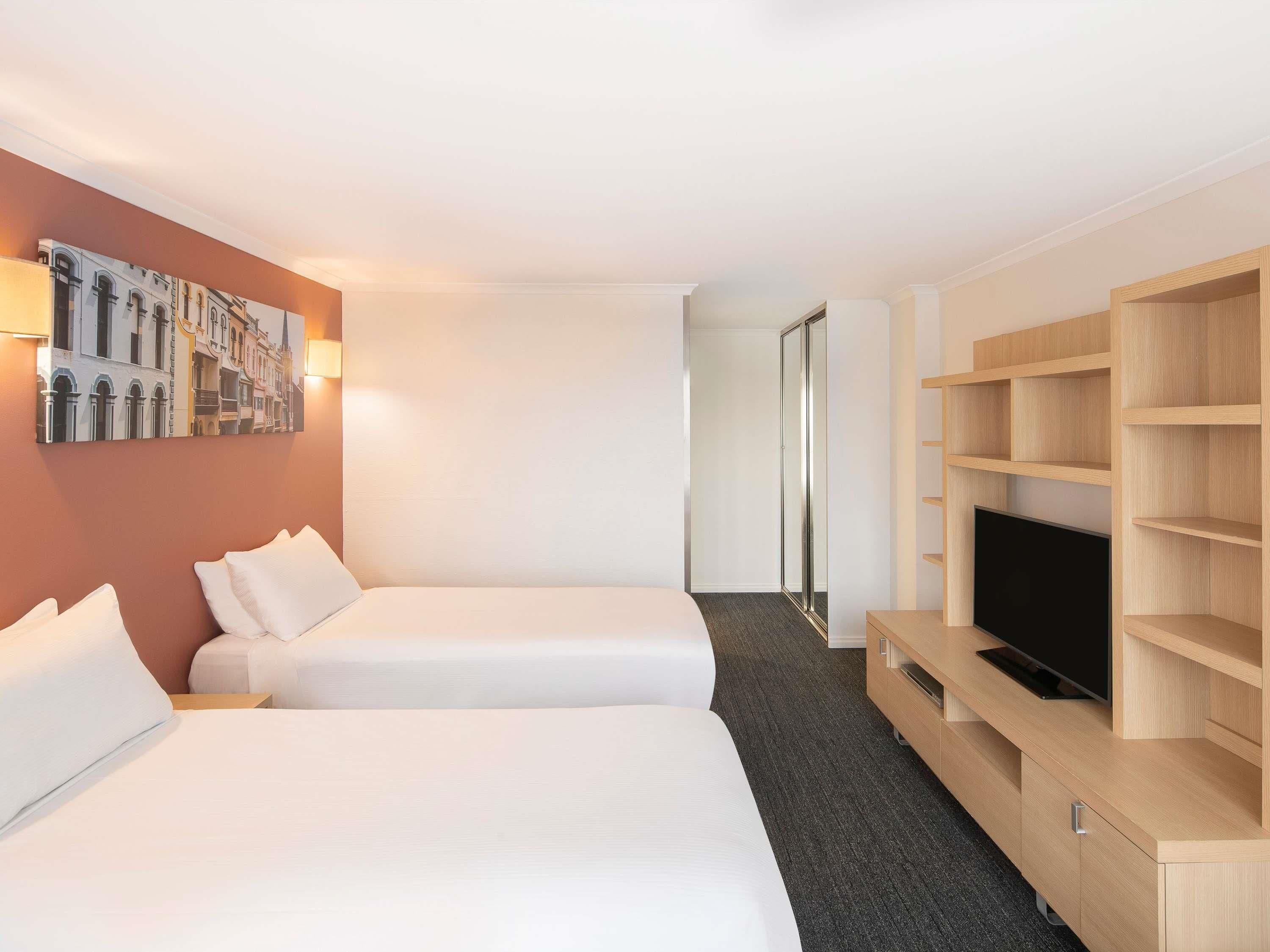 Quarto de hotel com duas camas, televisão e prateleira fixada na parede.