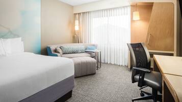 Quarto de hotel com cama, escrivaninha, cadeira, mesinha e janela com cortinas.