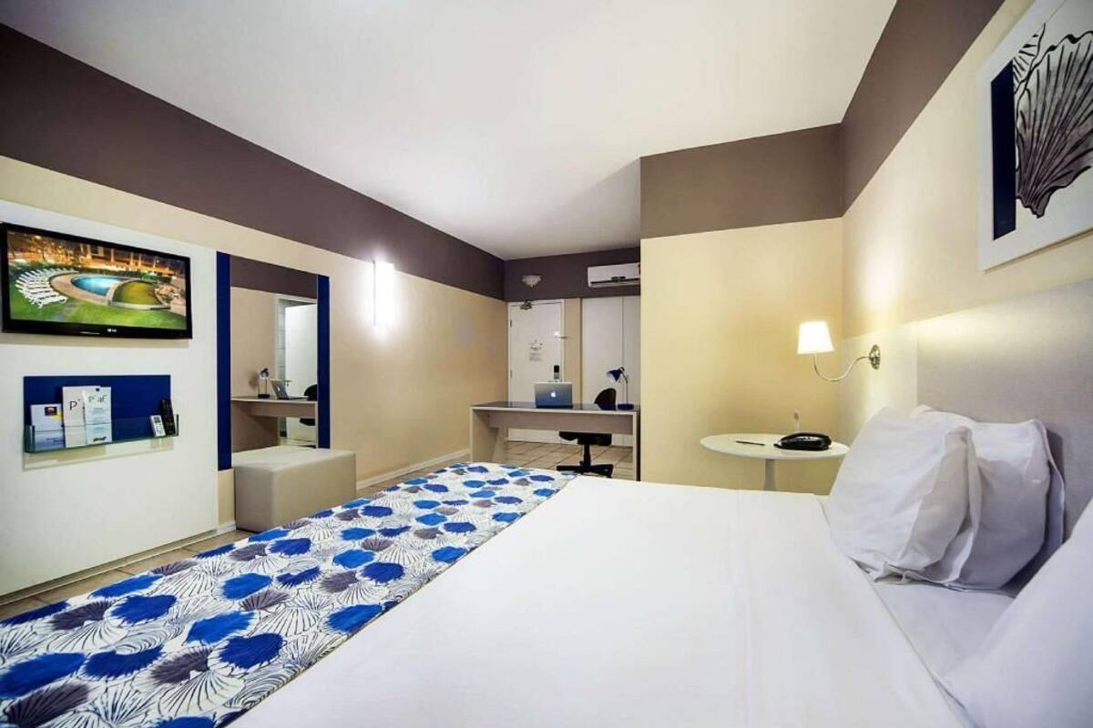 Quarto de hotel com cama, escrivaninha, cadeira, mesa pequena, televisão e espelho de banheiro.