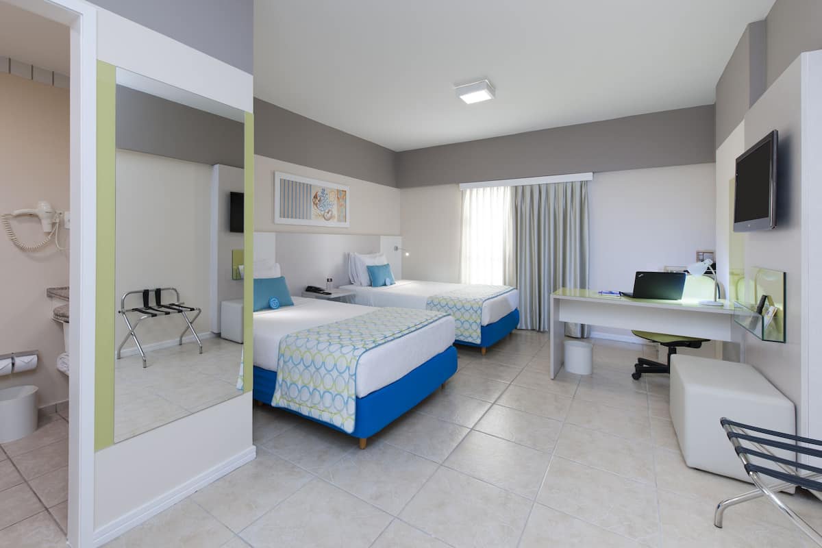 Quarto de hotel com duas camas, uma escrivaninha, uma cadeira, televisão e banheiro.