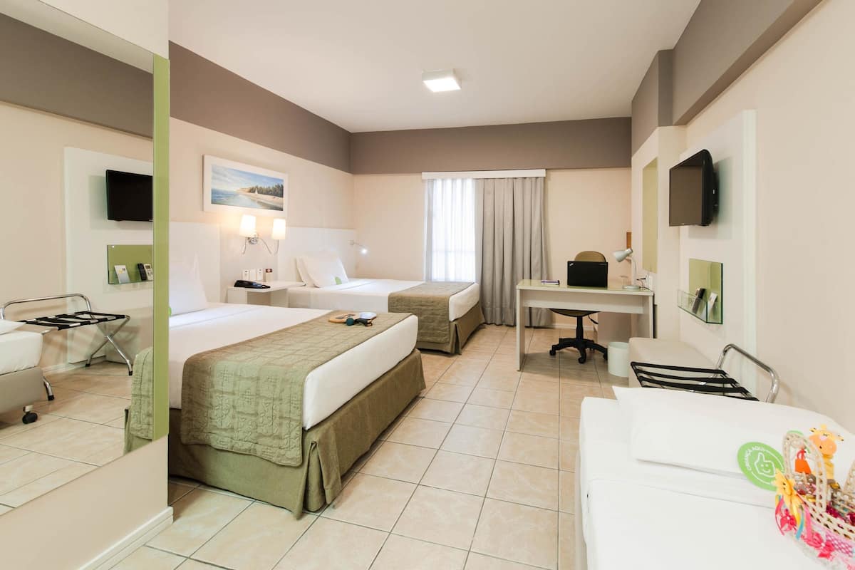 Quarto de hotel com duas camas, uma escrivaninha, uma cadeira, televisão e banheiro com chuveiro e vaso sanitário.