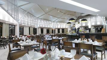 Uma ampla sala de jantar com várias mesas preparadas para o serviço, grandes janelas e um teto alto com iluminação natural.