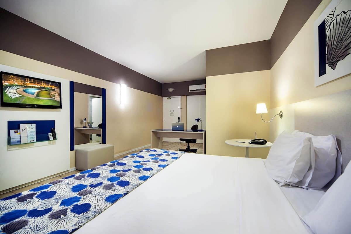 Quarto de hotel com cama, escrivaninha, cadeira, televisão, espelho, abajur e mesa pequena.