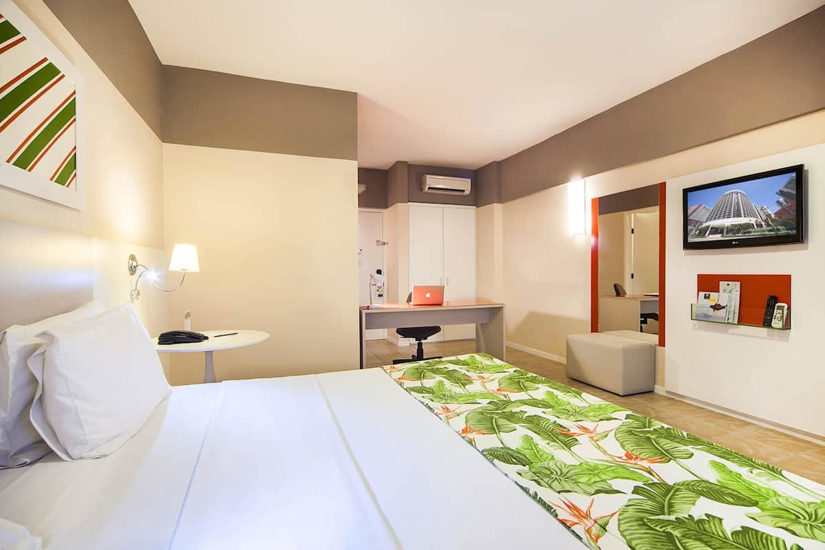 Quarto de hotel com uma cama, escrivaninha, cadeira, televisão e pequena mesa com abajur.