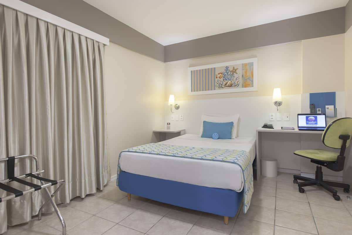 Quarto de hotel com cama, escrivaninha, cadeira e cortinas.