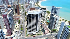 Um edifício alto com fachada curva, rodeado por outros edifícios altos, uma praia e um céu azul límpido.