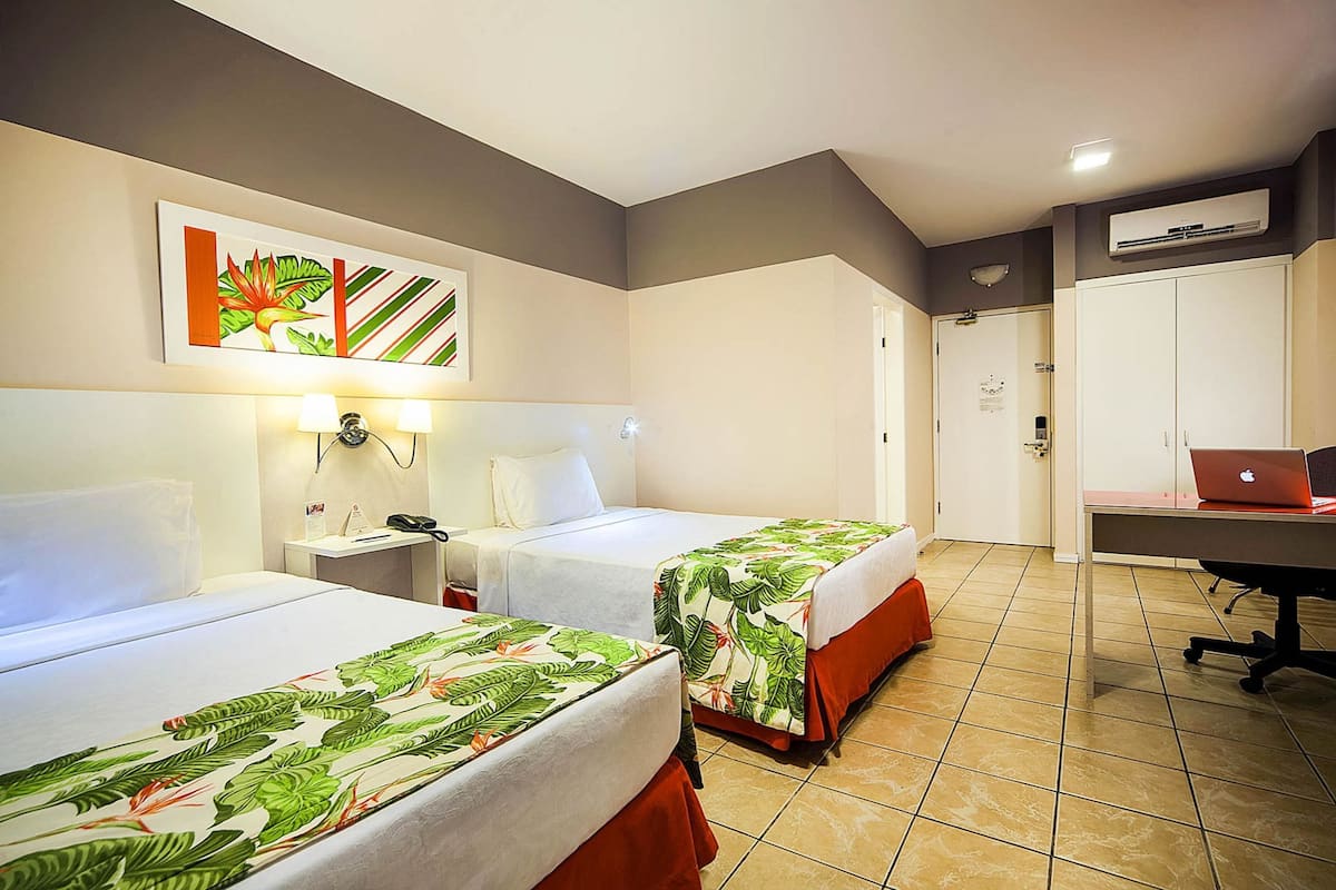 Quarto de hotel com duas camas, uma secretária com um computador portátil e uma porta que dá para outro quarto.