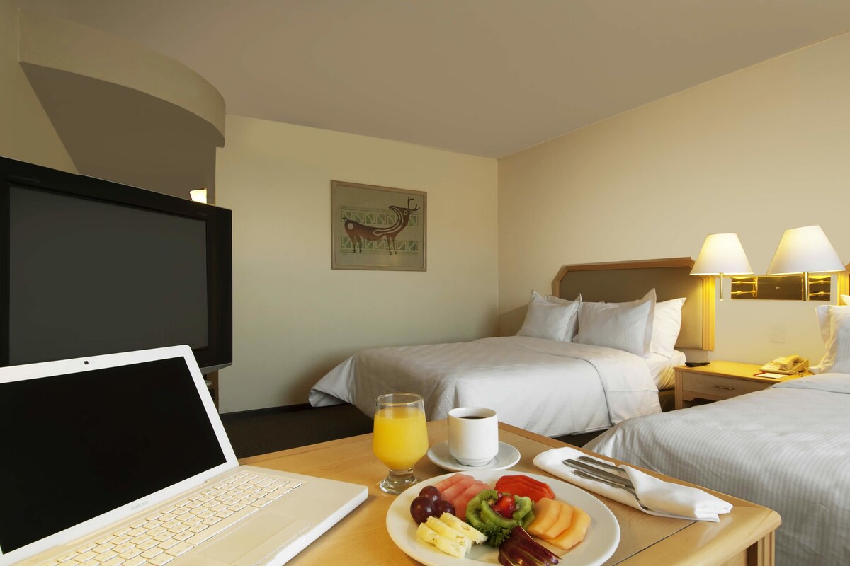 Quarto de hotel com cama, mesa de cabeceira, luminária e um laptop sobre a escrivaninha.