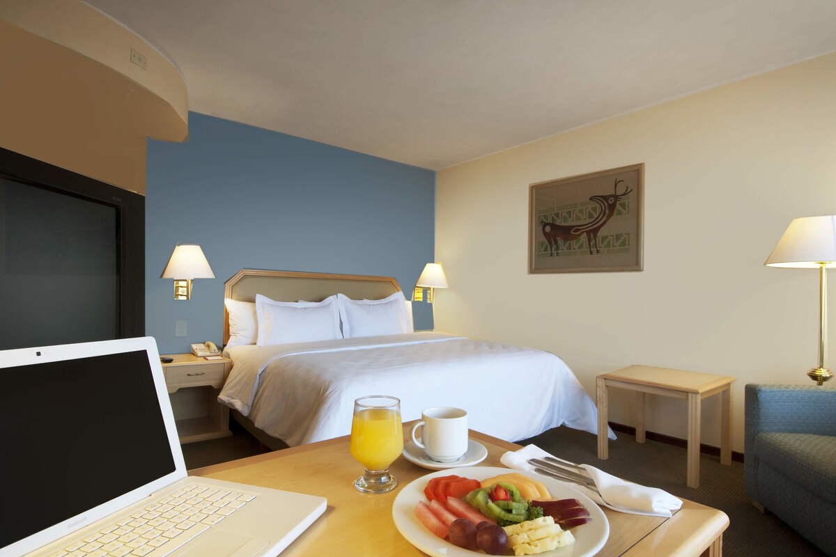 Quarto de hotel com cama, mesas de cabeceira, um laptop, pequeno-almoço com fruta e sumo, e um quadro na parede.