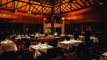 Restaurante com iluminação suave, vigas de madeira no teto, mesas com toalhas brancas e cadeiras.