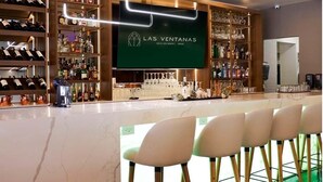 Uma área de bar com bancada de mármore, banquetas brancas e prateleiras com garrafas e copos.