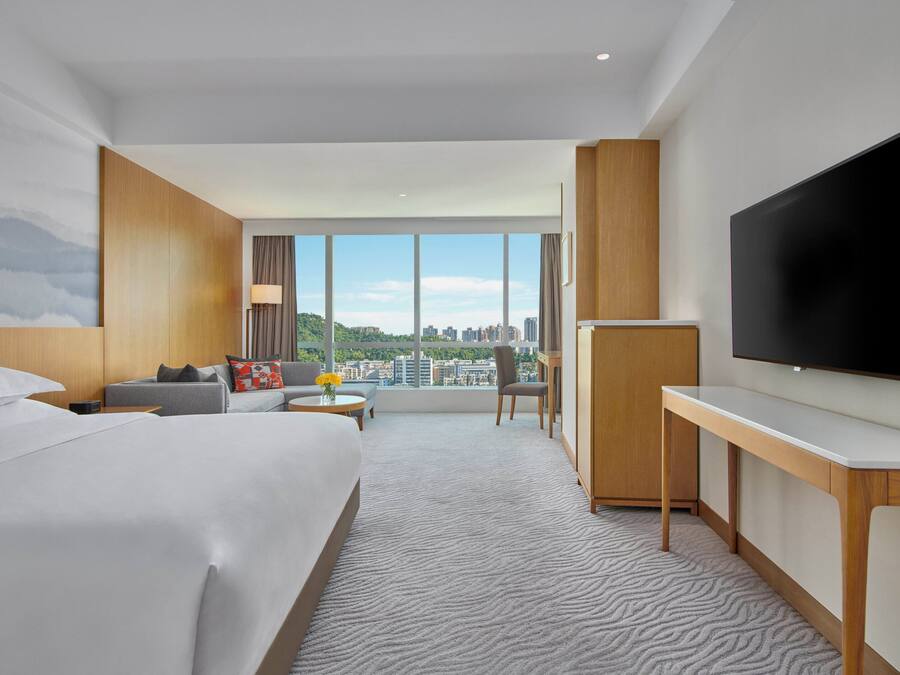 Quarto de hotel com cama, escrivaninha, televisão e uma grande janela com vista para a cidade.