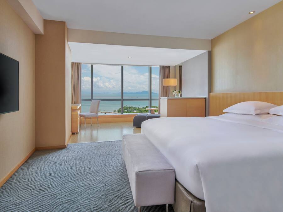 Quarto de hotel com uma cama grande, televisão de tela plana, área de estar com vista para o mar e uma pequena mesa com luminária.
