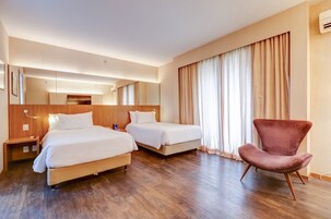Quarto de hotel com duas camas, cabeceira de madeira, um espelho grande, uma cadeira e uma janela com cortinas.