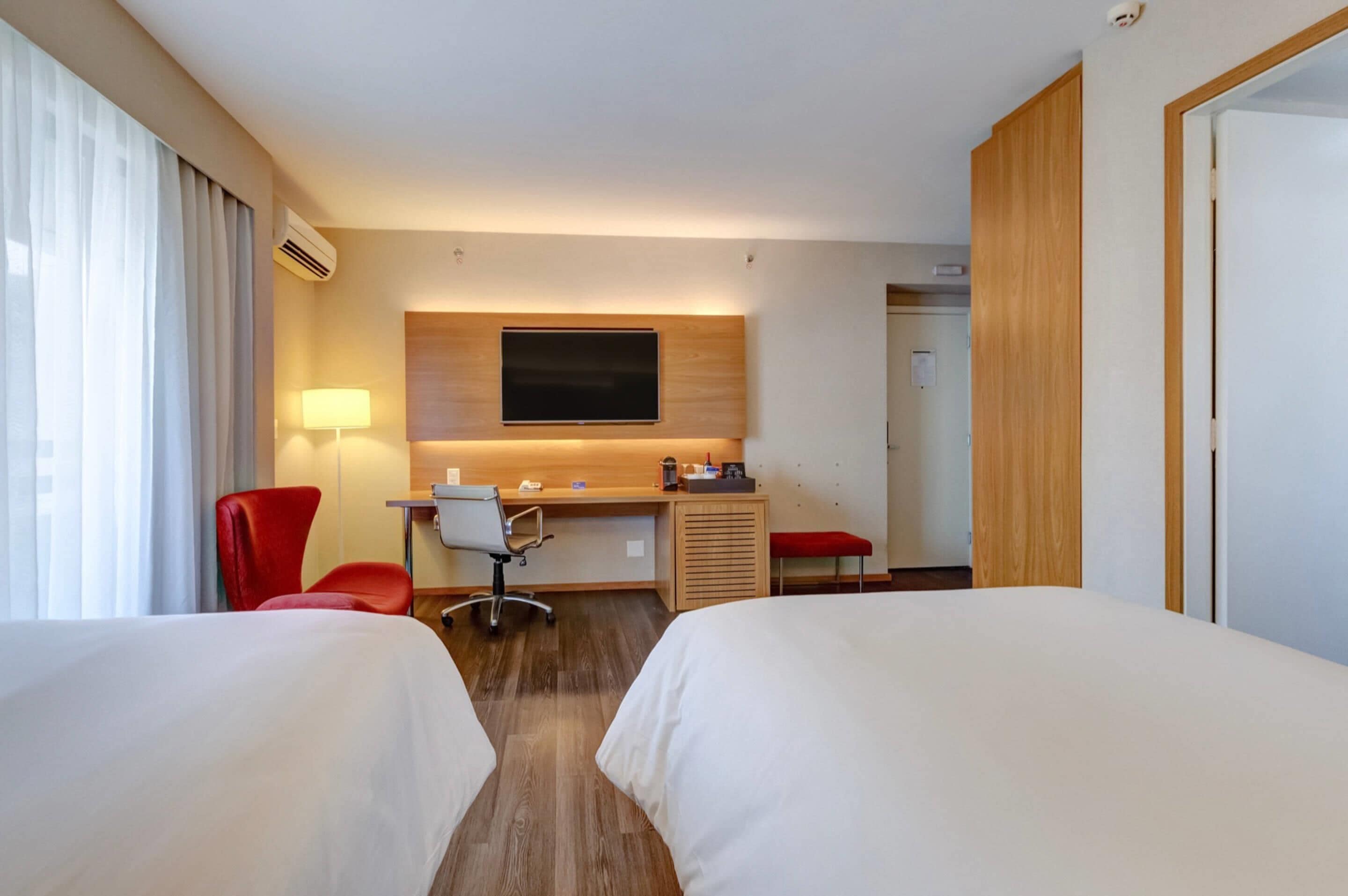 Quarto de hotel com duas camas, uma mesa com cadeira, uma TV e um abajur.