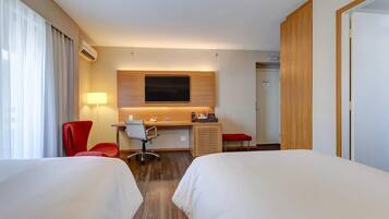 Quarto de hotel com duas camas, uma mesa com cadeira, uma TV e um abajur.