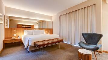 Quarto de hotel com uma cama grande, um banco de madeira, uma cadeira e uma pequena mesa redonda.