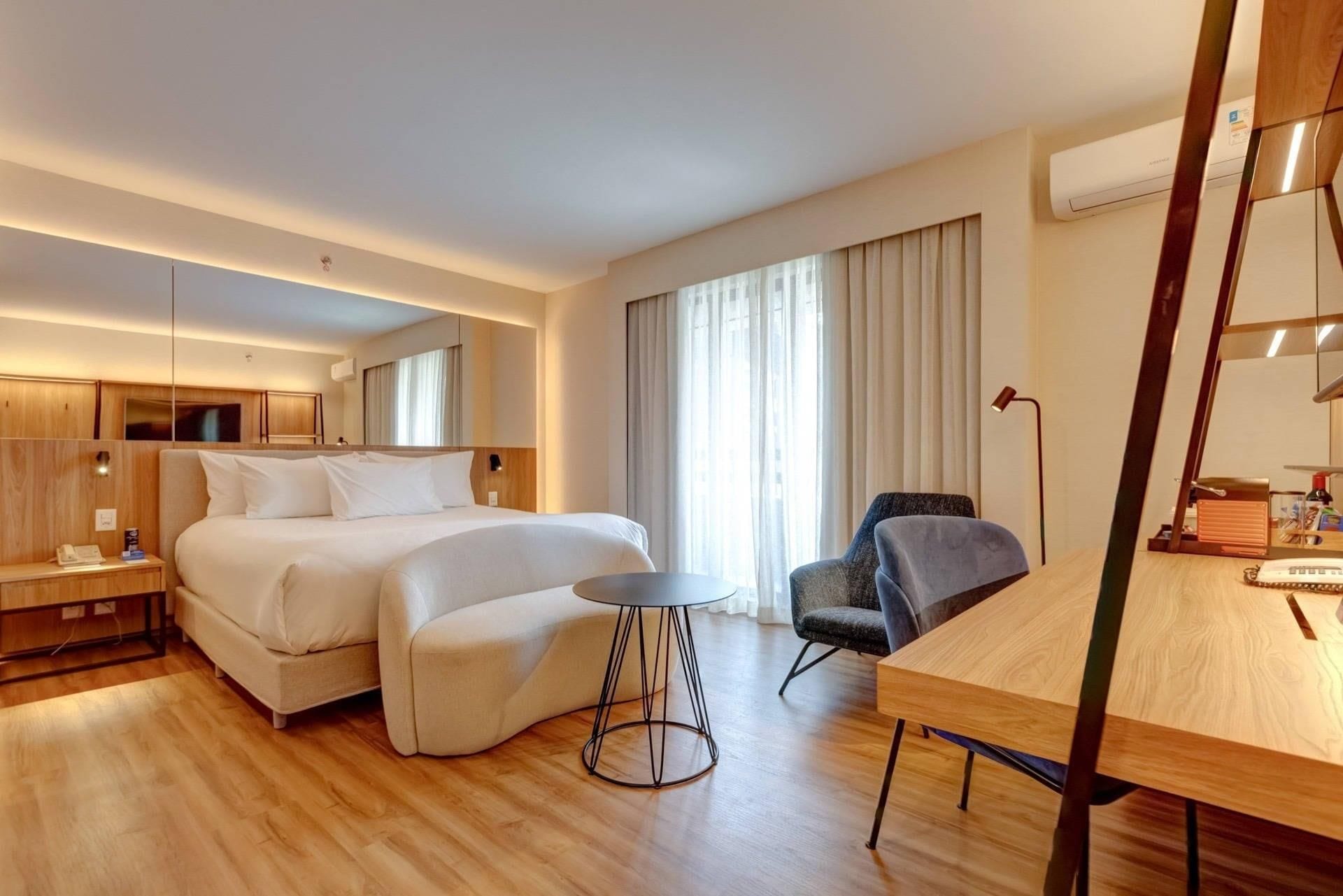 Quarto de hotel com uma cama grande, uma escrivaninha, uma cadeira, uma mesinha e um espelho.