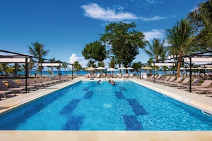 Piscina com espreguiçadeiras e guarda-sóis, rodeada por palmeiras e um céu azul claro.