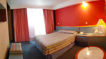 Quarto de hotel com cama, mesa de cabeceira com abajur, um banquinho e janela com cortinas.