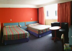 Quarto de hotel com duas camas, uma mesinha, uma cadeira e janela com cortinas.