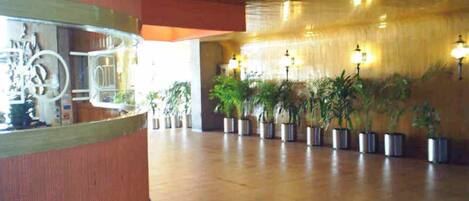 Um hall amplo com piso lustroso, uma mesa de recepção e uma fileira de plantas em vasos.