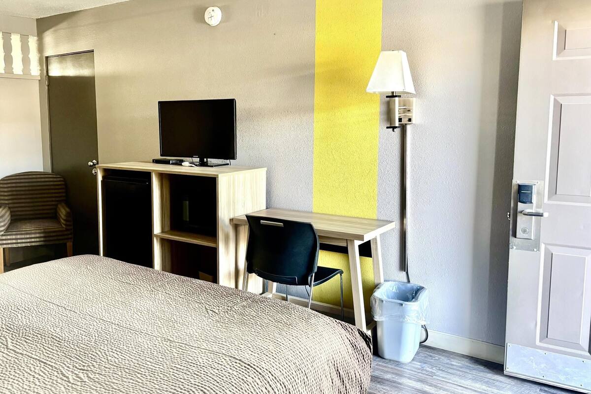 Quarto de hotel com cama, escrivaninha com televisão, cadeira, abajur e lixeira.
