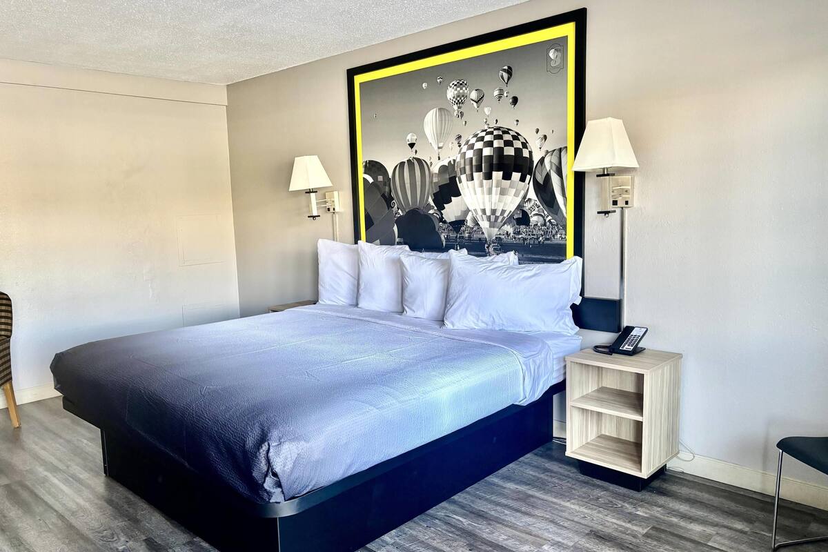 Quarto de hotel com cama, luminárias de cabeceira, mesa de cabeceira e uma obra de arte na parede.