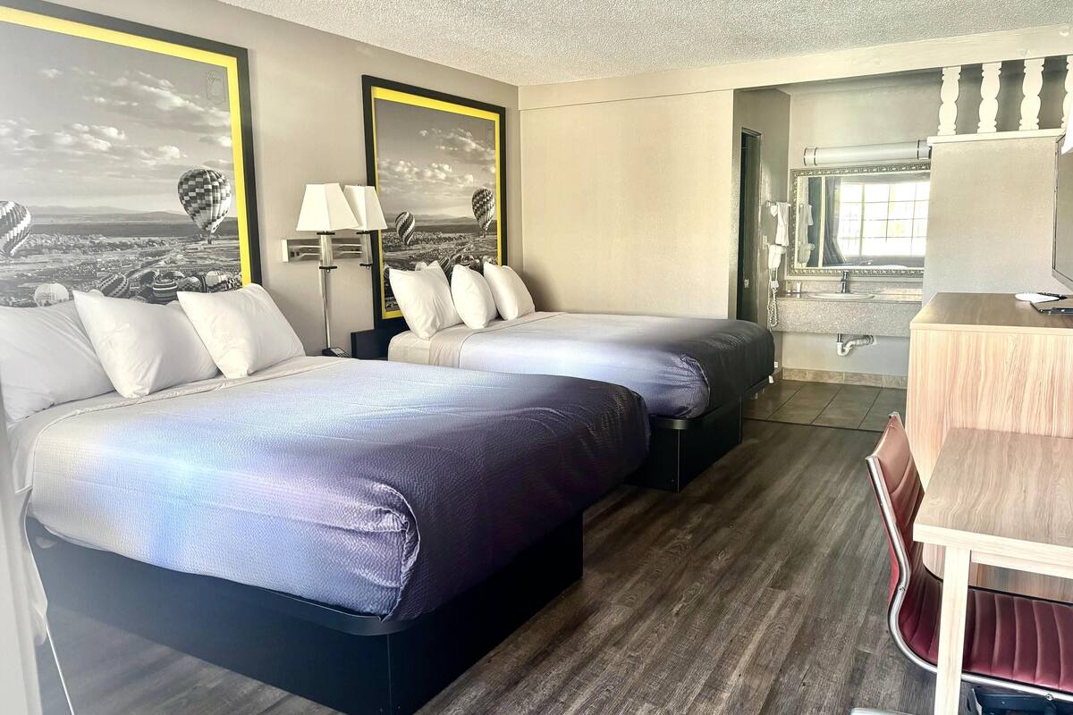 Quarto de hotel com duas camas, uma escrivaninha, um espelho e uma janela.