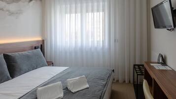 Quarto de hotel com cama, escrivaninha, televisão e janela com cortinas leves.