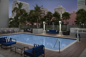 Piscina com espreguiçadeiras e palmeiras, cercada por edifícios.
