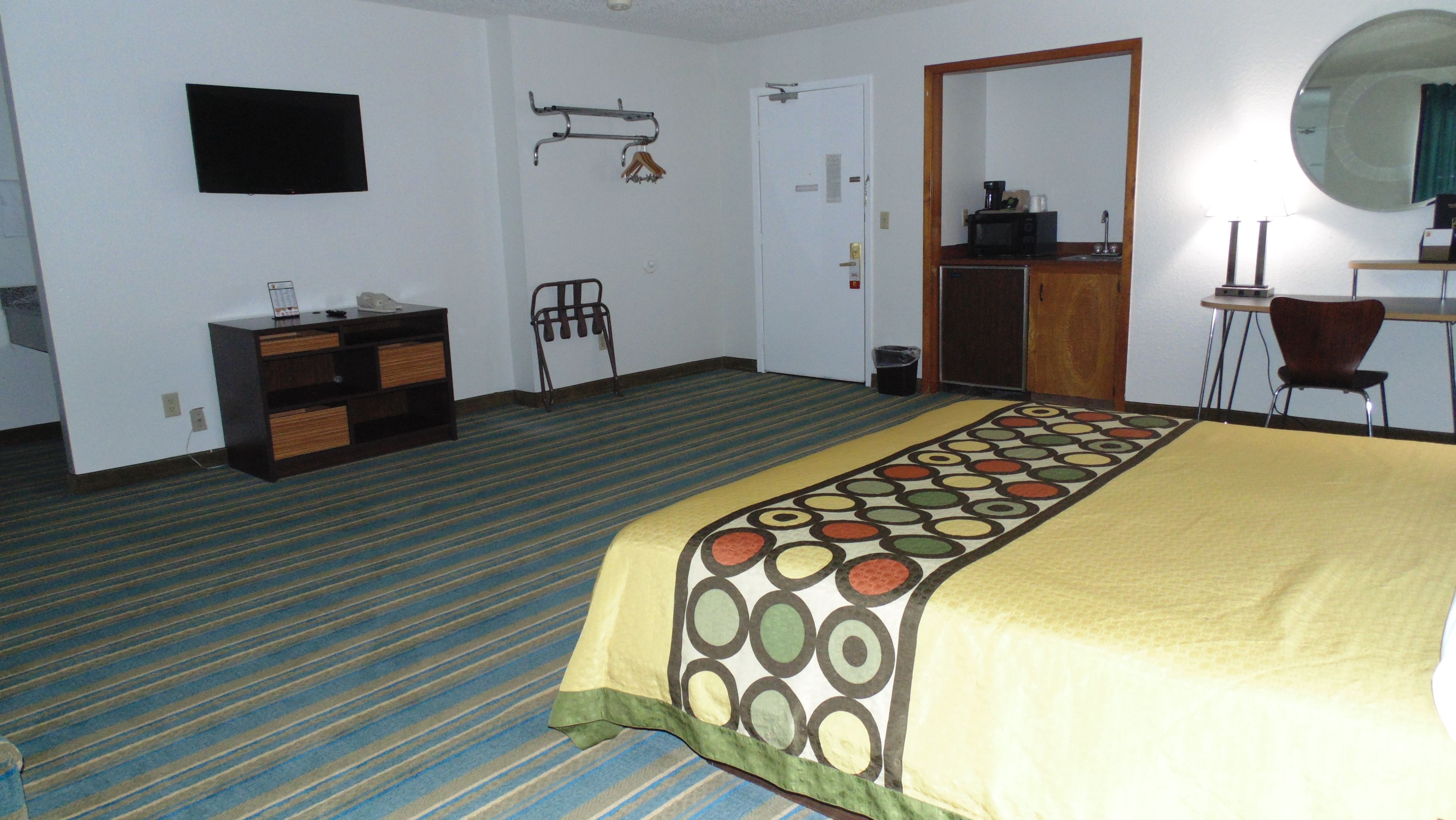 Quarto de hotel com uma cama, uma escrivaninha, uma cadeira, uma televisão, um cabideiro e uma porta.